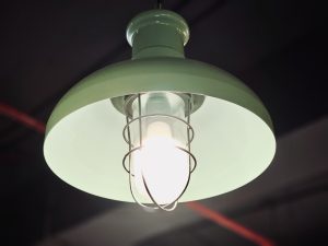 A green metal hanging lamp glowing softly indoors, creating a calm and warm mood. The simple design and focused light highlight everyday interior details, captured in Kozhikode, Kerala, with a clean and minimal background.  